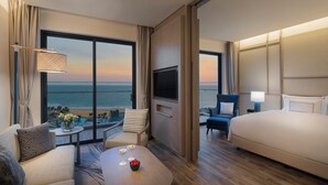 Quarto de hotel com uma janela grande, televisão, cama, sofá, uma mesinha com uma tigela de fruta e uma cadeira.