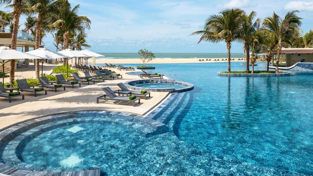 Área da piscina com espreguiçadeiras e guarda-sóis, uma banheira de hidromassagem e vista para a praia.