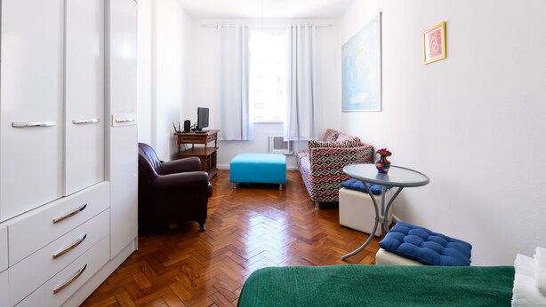 Quarto pequeno com guarda-roupa, sofá, mesa e janela com cortinas.