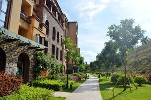 Caminho que leva a um edifício com fachada de tijolos e janelas em arco, rodeado por vegetação bem cuidada e uma paisagem acidentada.