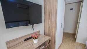 Quarto com televisão de tela plana na parede, escrivaninha de madeira com controle remoto e um vaso de flores, e porta que dá para outro cômodo.