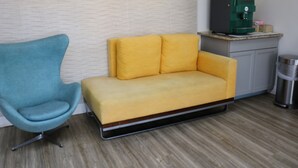 Um quarto com uma poltrona azul-turquesa, um sofá amarelo, um espaço de cozinha compacta com cafeteira e uma lixeira.