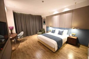 Quarto de hotel moderno com uma cama grande, uma escrivaninha com cadeira, uma mesinha com um vaso de flores e uma janela ampla com cortinas.