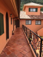 Um corredor com piso ladrilhado, corrimão de metal e uma porta que dá para um edifício com telhado de telhas.