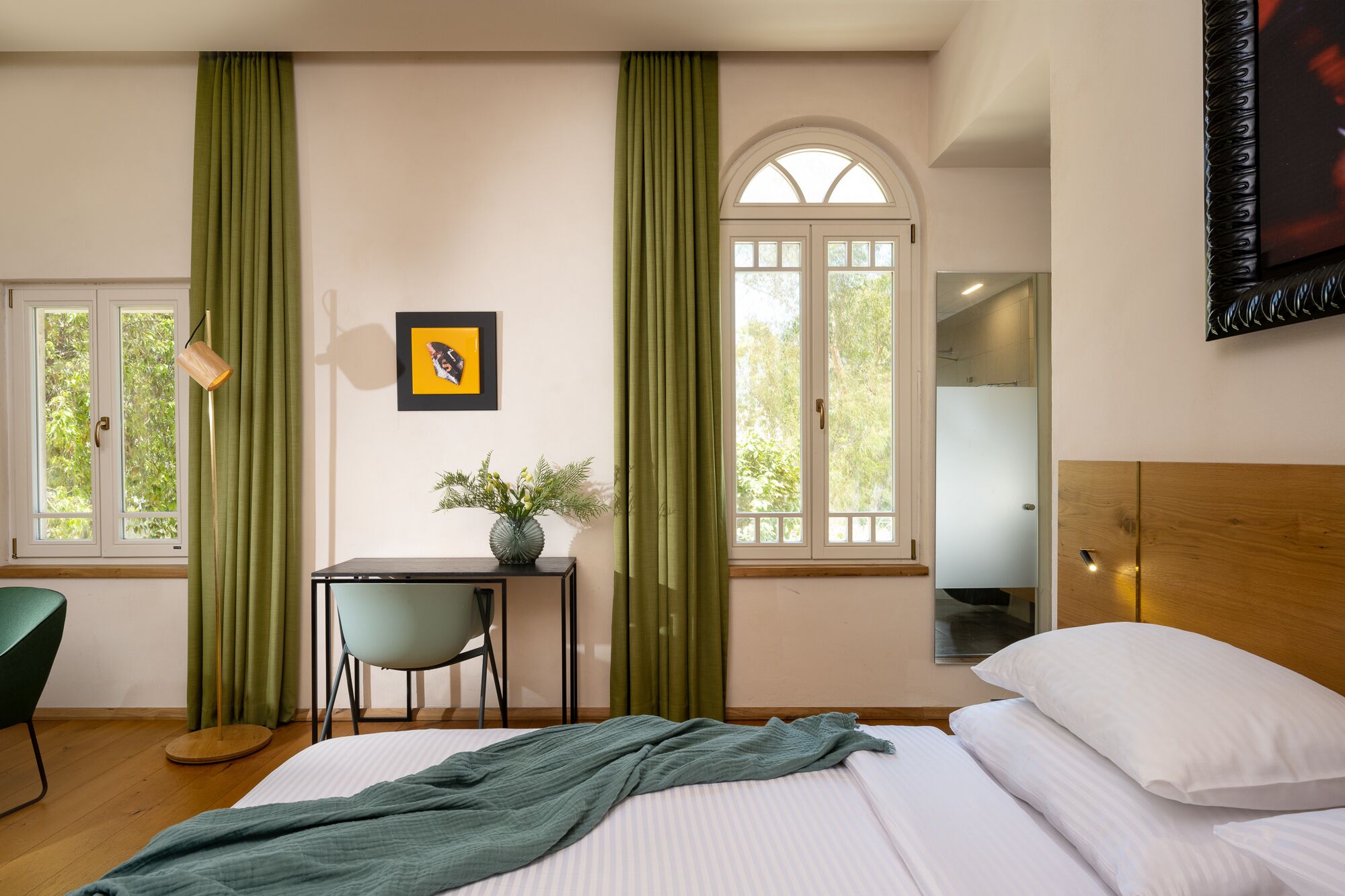 Quarto de hotel com cama, escrivaninha e cadeira, planta em vaso e janela com cortinas.