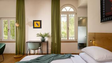 Quarto de hotel com cama, escrivaninha e cadeira, planta em vaso e janela com cortinas.