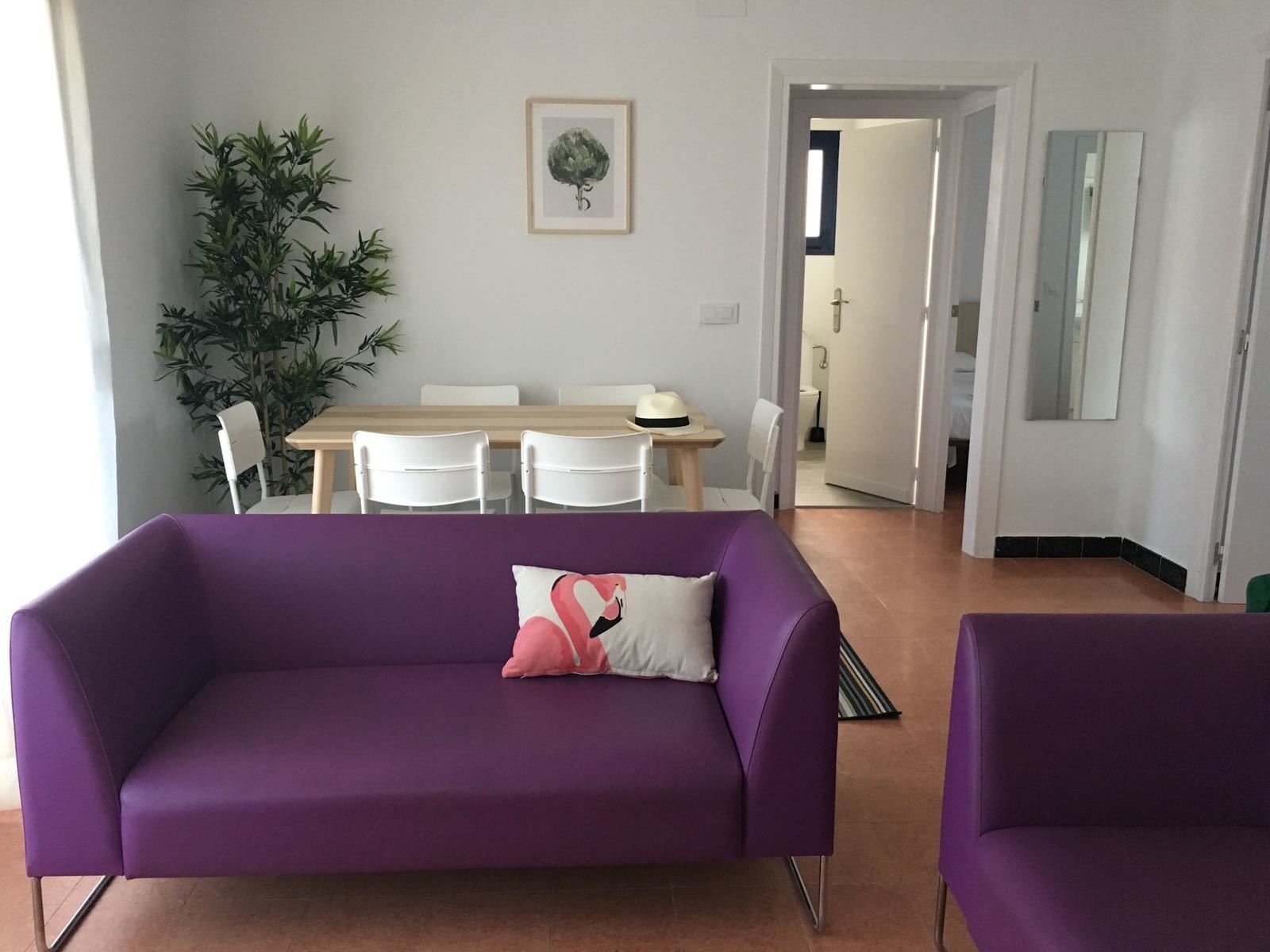 Uma sala de estar com sofá roxo, um espaço de jantar com mesa e cadeiras, e uma planta grande.