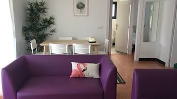 Uma sala de estar com sofá roxo, um espaço de jantar com mesa e cadeiras, e uma planta grande.