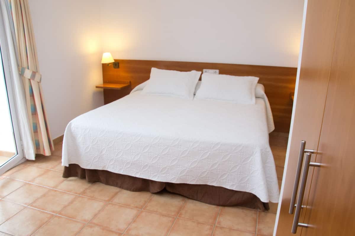 Uma cama bem arrumada com cabeceira de madeira, luminária ao lado e vista para o exterior através de uma janela com cortinas.