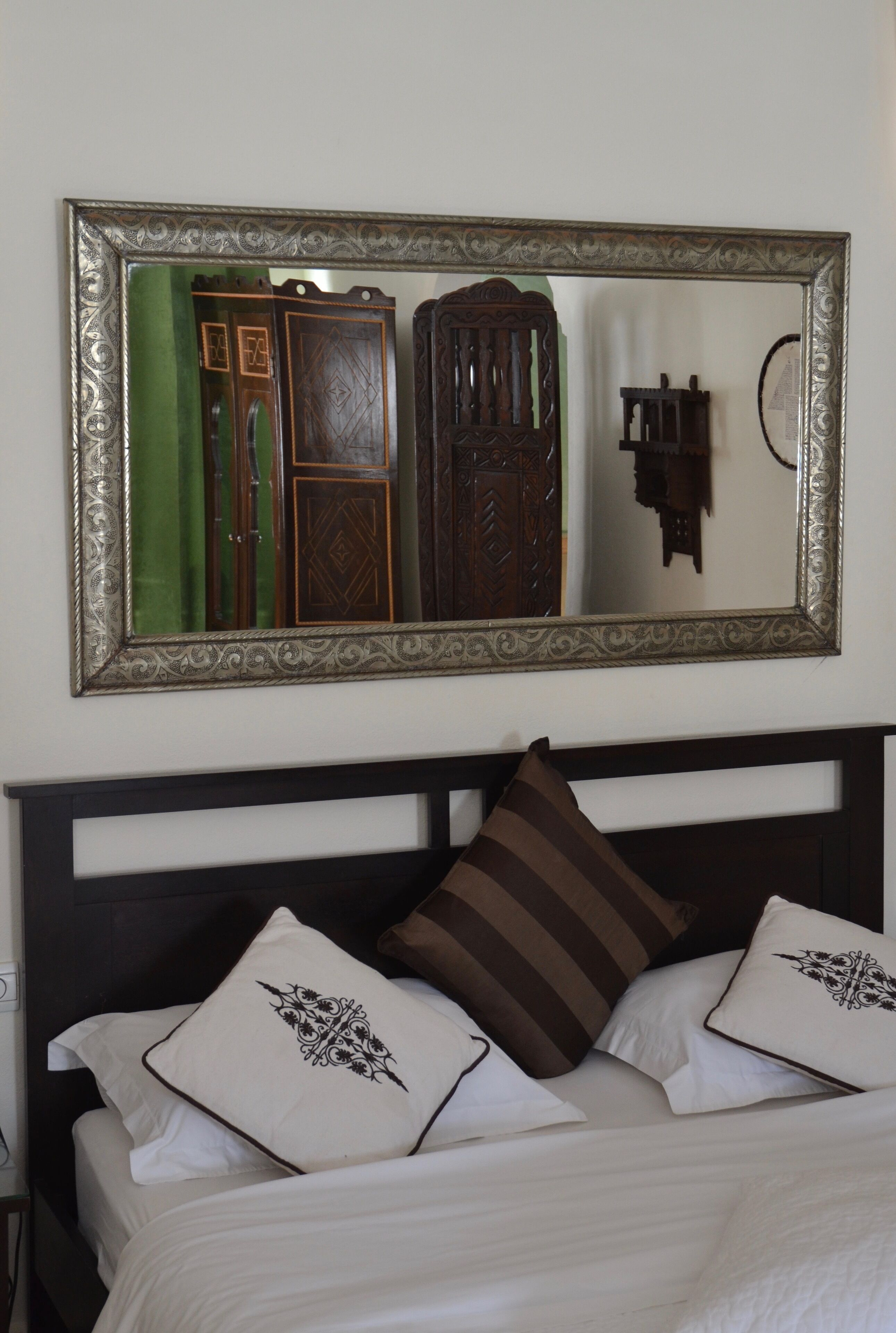 Cama com roupa de cama branca e almofadas decorativas, cabeceira escura e um espelho grande e ornamentado acima.