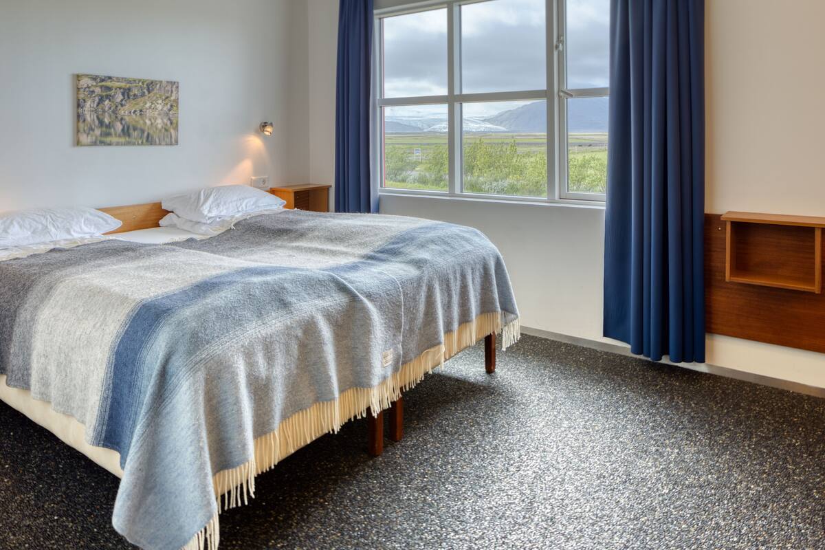 Um quarto com cama, mesa de cabeceira, janela com vista para o exterior e um quadro na parede.