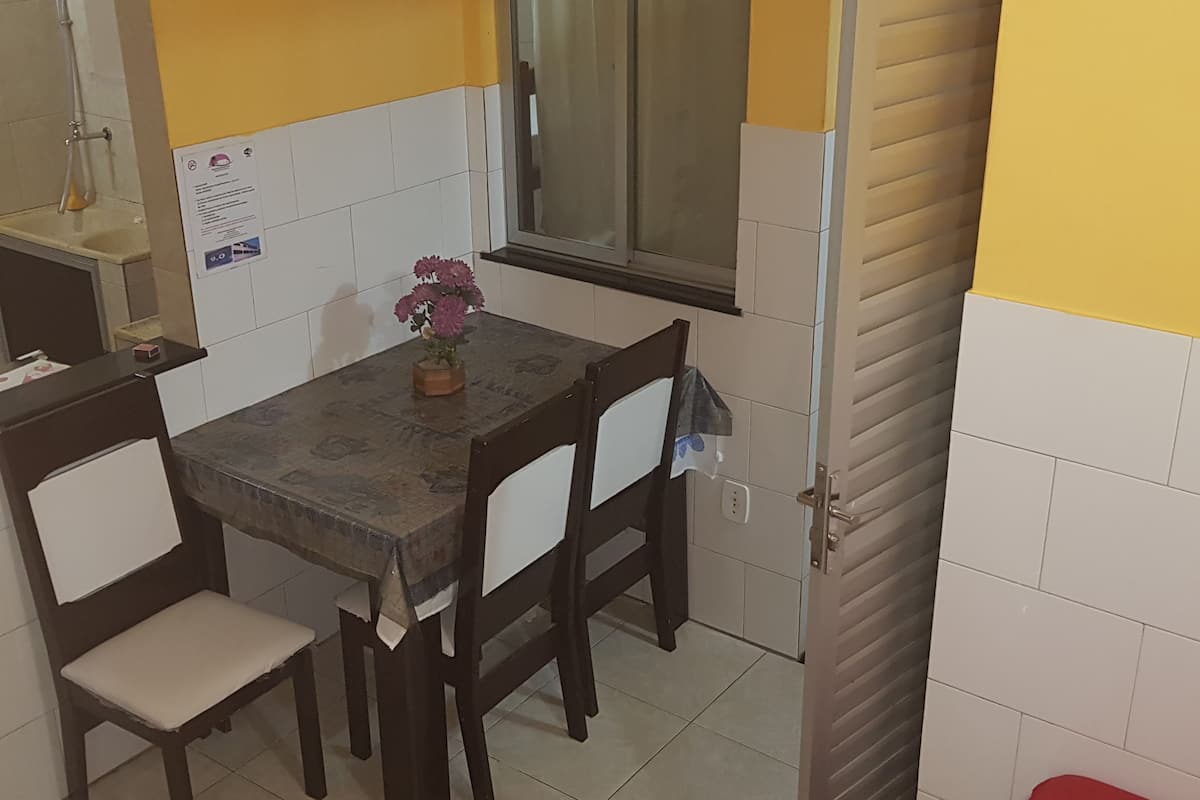 Uma área de refeições com uma mesa e quatro cadeiras, um vaso com flores, uma janela e uma porta.