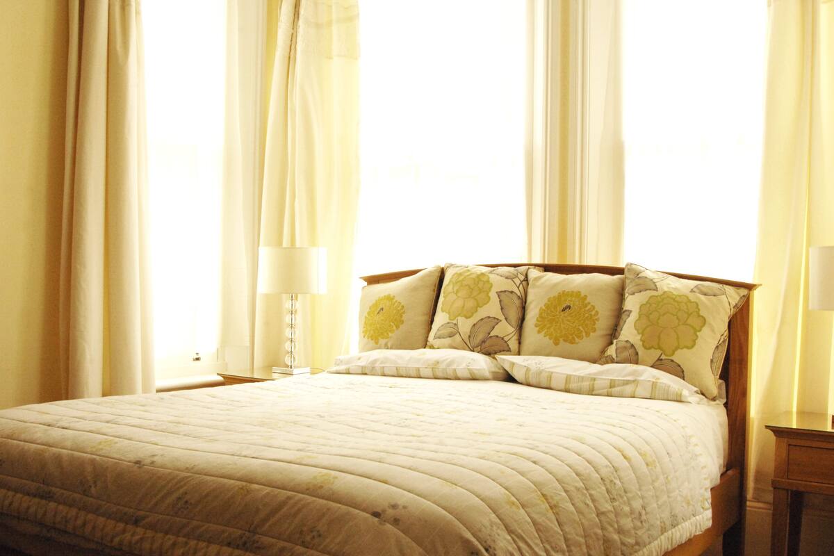 Uma cama bem arrumada com almofadas florais, uma luminária de cabeceira e uma janela com cortinas.