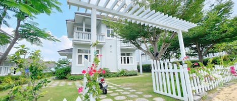 Uma casa branca de dois andares com varanda, cercada por um portão branco em estilo picket e um jardim com caminho de pedra trabalhado.