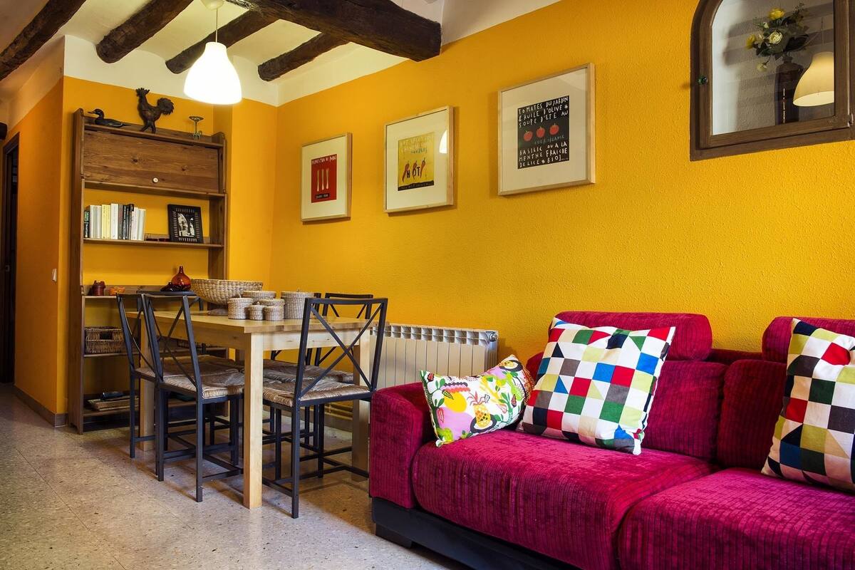 Um quarto aconchegante com uma mesa e cadeiras de madeira, um sofá vermelho com almofadas coloridas e uma estante repleta de livros e objetos decorativos.