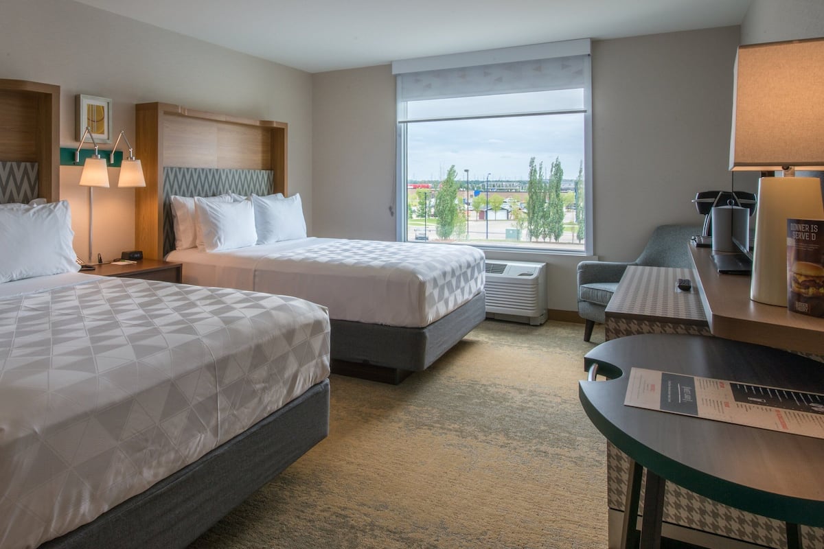 Quarto de hotel com duas camas, uma escrivaninha, uma cadeira e janela com vista para edifícios.