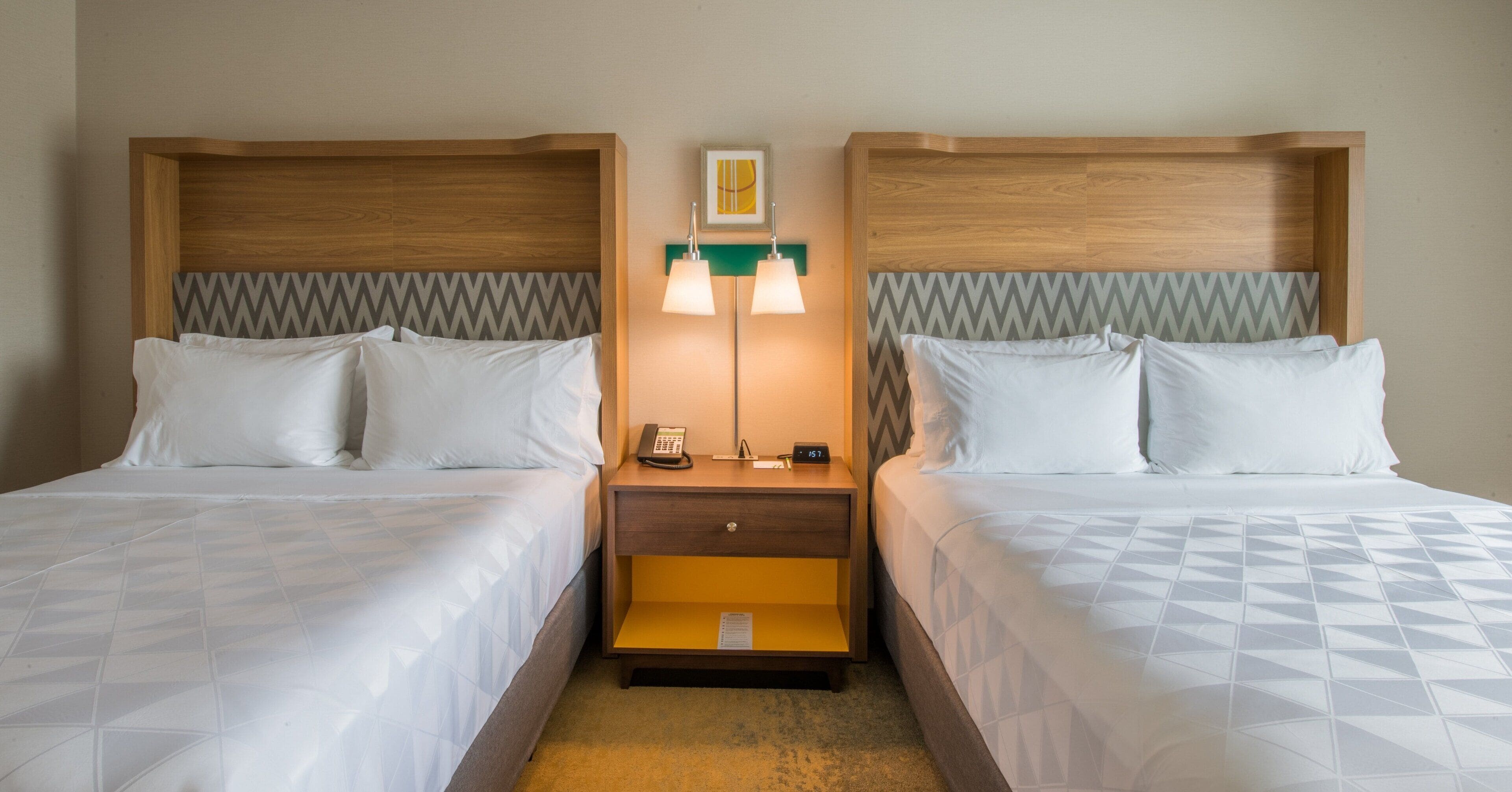 Quarto de hotel com duas camas, cada uma com cabeceira de madeira, mesa de cabeceira com abajur e uma pequena mesa entre as camas.
