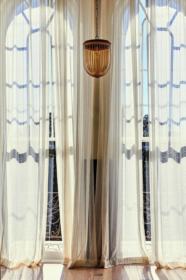 Quarto com cortinas leves, luminária pendente e vista pela janela.