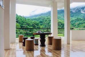 Quarto com chão de madeira, uma mesa redonda e vários bancos. Possui uma varanda com vista para uma paisagem verdejante e montanhas ao longe.