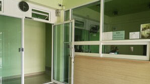 Área de recepção de hotel com porta de vidro, relógio e balcão de atendimento com computador e planta.
