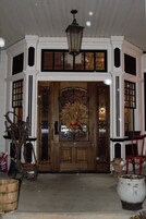 Uma porta de madeira com uma guirlanda decorativa, um candeeiro em cima e uma cadeira vermelha à direita.