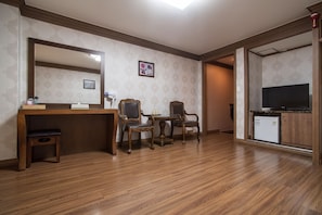 Quarto com uma escrivaninha de madeira, duas cadeiras, um pequeno frigorífico, televisão e um espelho.