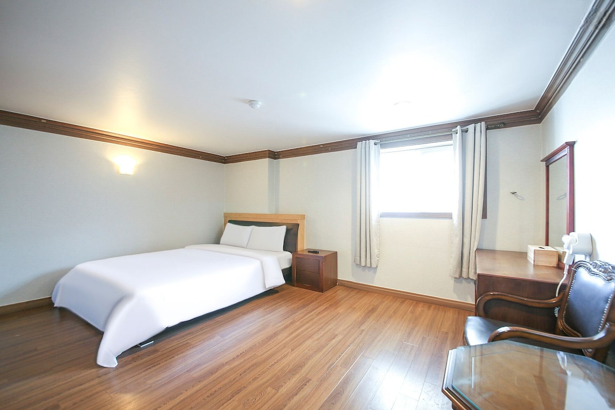 Quarto de hotel com cama, mesa de cabeceira, cadeira e uma janela grande com cortinas.