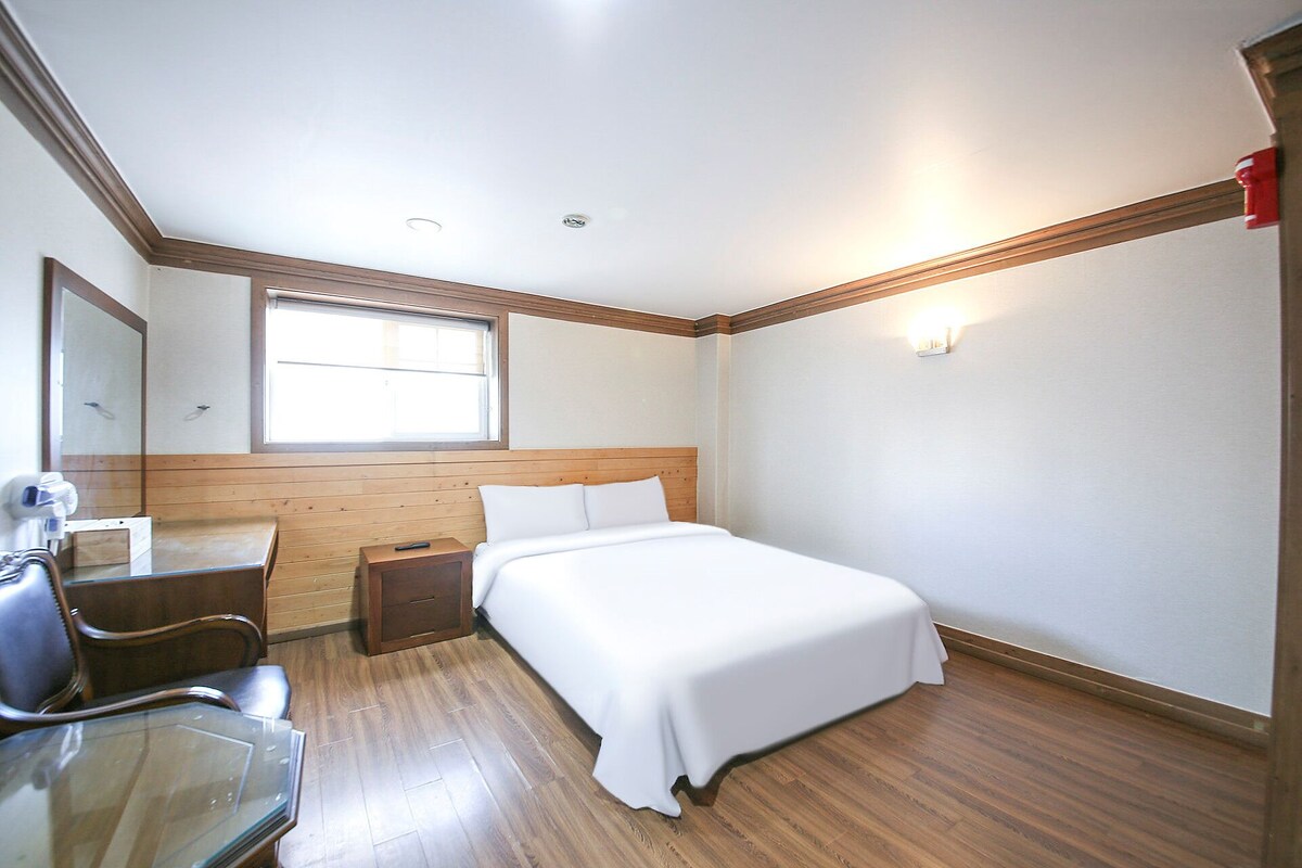Quarto de hotel com cabeceira de madeira, uma cama de casal, uma pequena escrivaninha com cadeira, um espelho e uma janela.