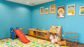 Uma área de recreação infantil com um escorrega vermelho, uma estrutura de escalada azul e uma estante de madeira com livros e brinquedos.