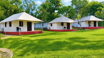 Três pequenas cabanas térreas, com base vermelha e seções superiores brancas, cada uma com porta e janela, situadas em uma área gramada com árvores ao fundo.