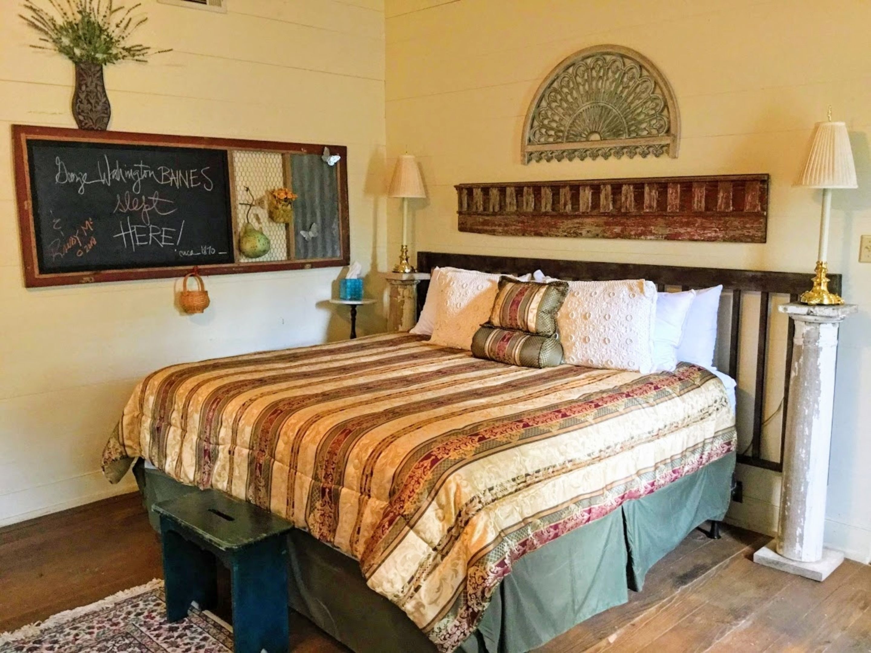 Cama bem arrumada com colcha estampada, mesa de cabeceira com abajur e um objeto decorativo fixado na parede acima da cama.