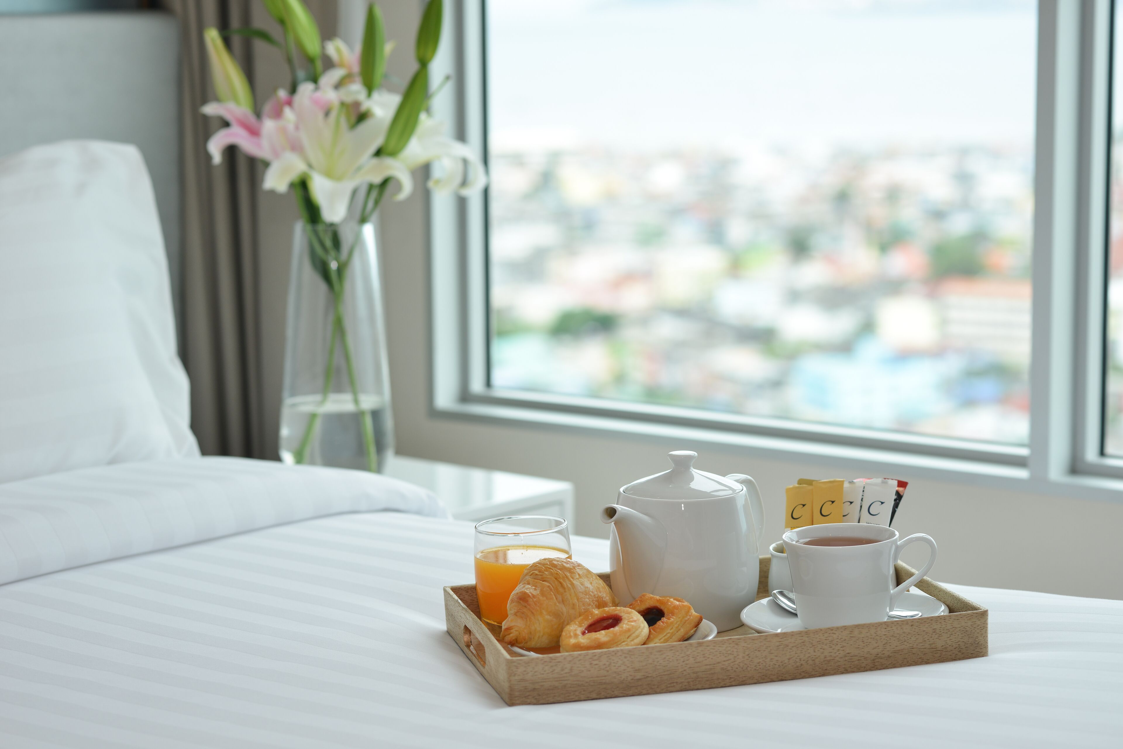 Bandeja de café da manhã bem arrumada com croissants, uma xícara de café e um copo de suco de laranja em uma cama, com vista para o horizonte urbano através de uma janela.