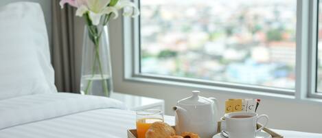 Bandeja de café da manhã bem arrumada com croissants, uma xícara de café e um copo de suco de laranja em uma cama, com vista para o horizonte urbano através de uma janela.