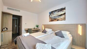 Quarto de hotel com uma cama grande, uma escrivaninha, uma cadeira, um quadro de uma cena de praia e uma porta que dá para outro quarto.