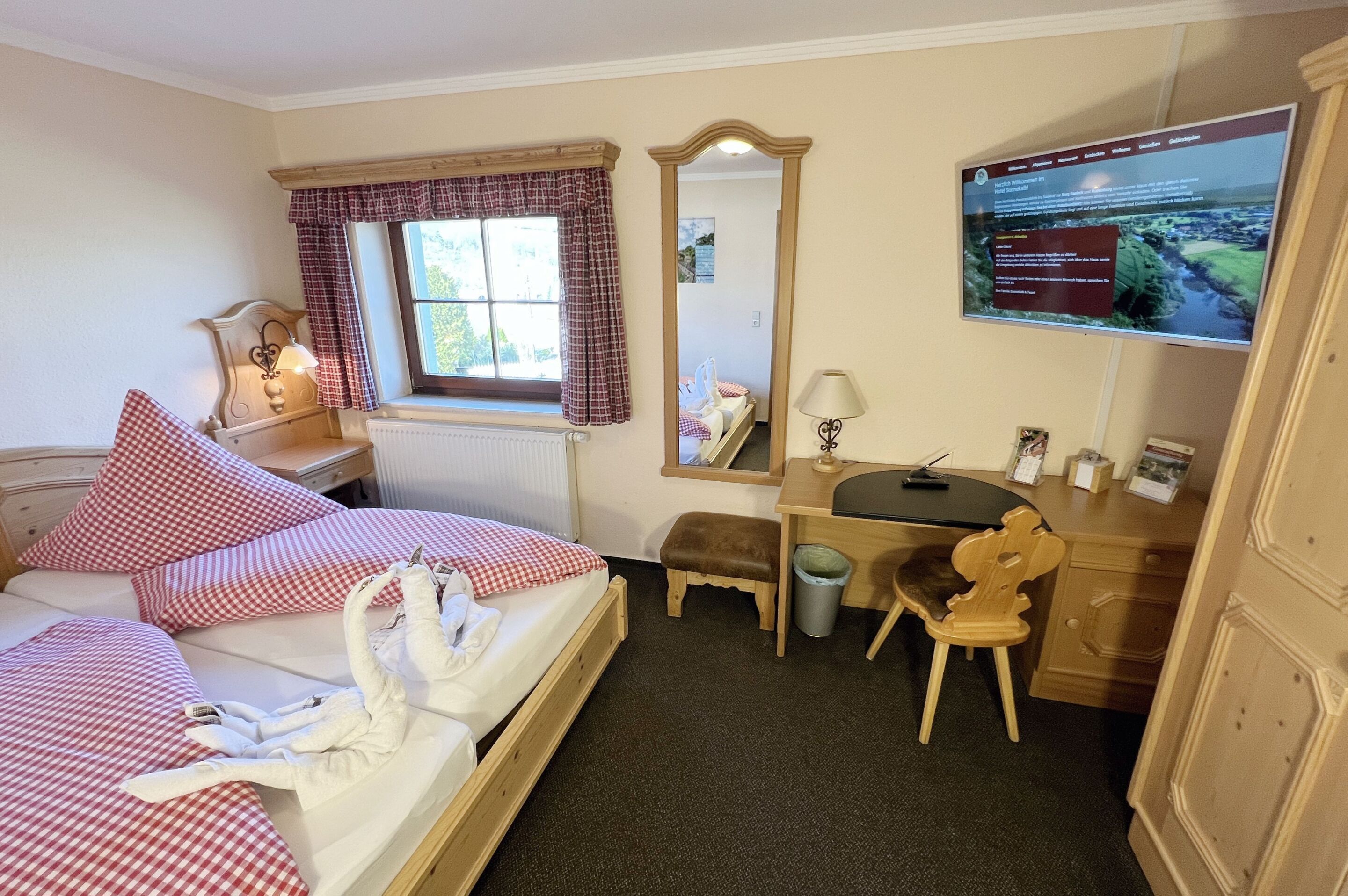 Quarto de hotel com duas camas, uma escrivaninha, uma cadeira, um espelho, uma janela com cortinas, um radiador e uma televisão.