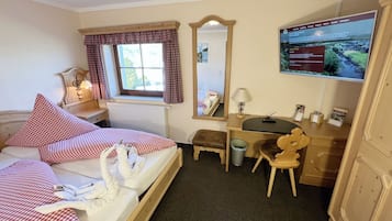 Quarto de hotel com duas camas, uma escrivaninha, uma cadeira, um espelho, uma janela com cortinas, um radiador e uma televisão.