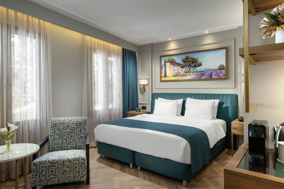 Quarto de hotel com cama grande, uma cadeira, mesa pequena com um vaso e um quadro na parede.