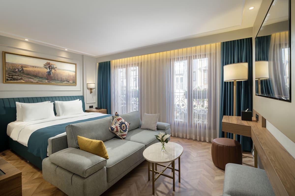 Quarto de hotel moderno com uma cama grande, um sofá cinzento, uma mesinha redonda pequena e uma escrivaninha de madeira.