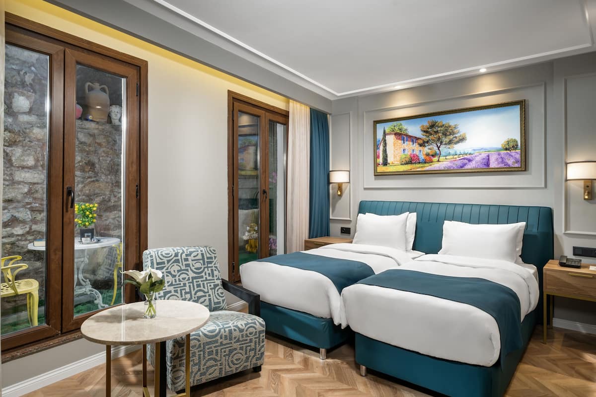 Quarto de hotel com cama, duas mesas de cabeceira, uma cadeira, uma mesinha e um quadro na parede.