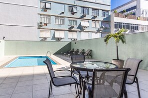 Uma área de piscina no terraço com mesa e cadeiras de vidro, uma palmeira e um edifício com várias varandas ao fundo.