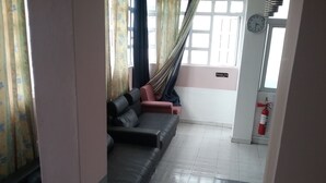 Quarto com sofá preto, poltrona rosa, janela com cortinas, relógio e extintor de incêndio.