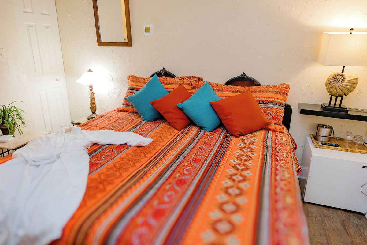 Cama bem arrumada com roupa de cama estampada e duas almofadas, mesa de cabeceira com luminária e prateleira pequena com objetos decorativos.