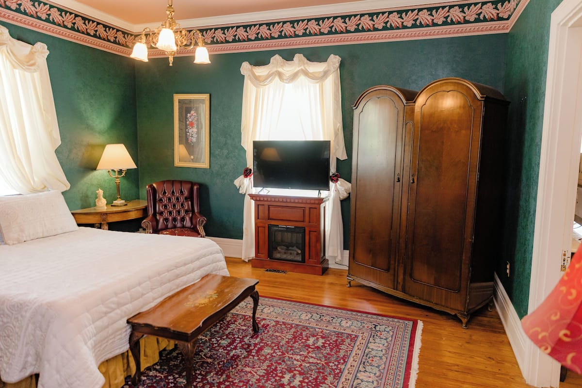 Um quarto com uma cama, uma cadeira de madeira, uma televisão, um guarda-roupa e uma lareira.