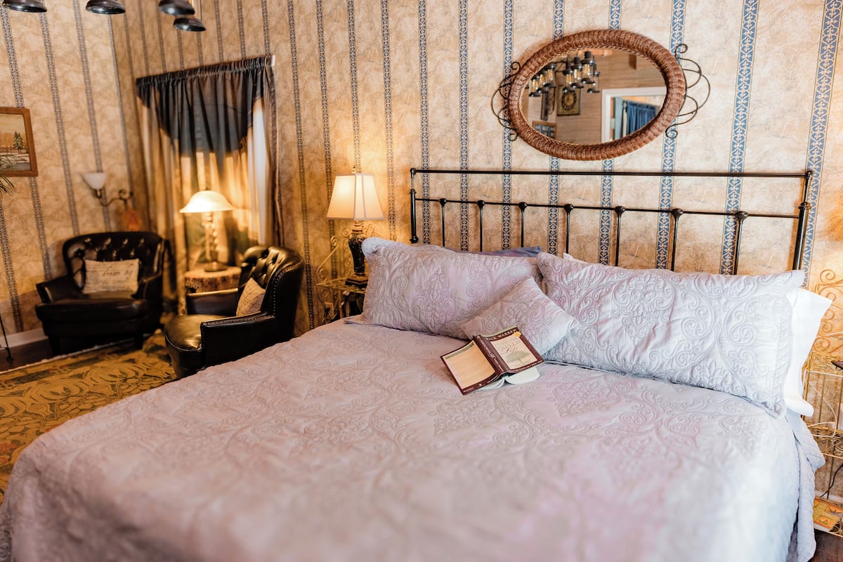 Uma cama bem arrumada com colcha estampada e travesseiros, um abajur, uma cadeira e um espelho na parede.