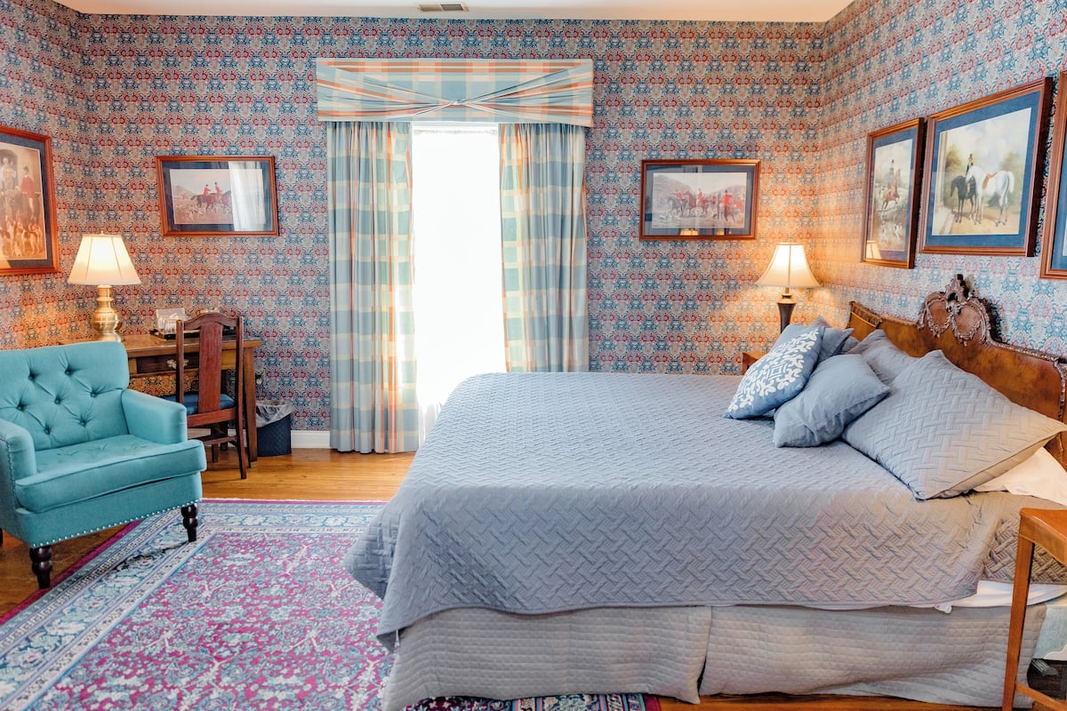Quarto com uma cama grande, duas luminárias, uma poltrona azul, uma escrivaninha de madeira e papel de parede estampado.