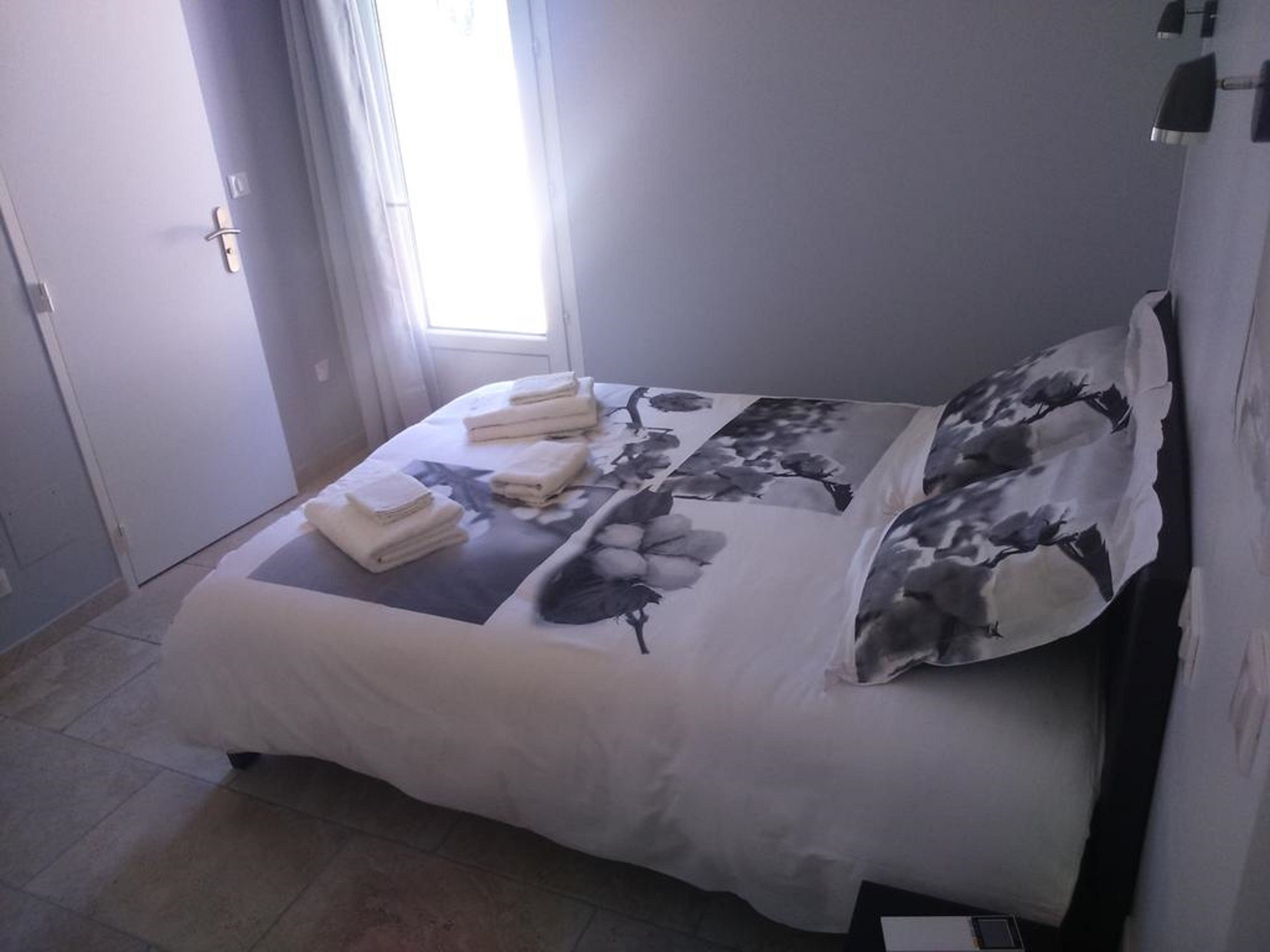 Uma cama bem arrumada com colcha e almofadas florais, uma janela com cortinas e uma porta à esquerda.