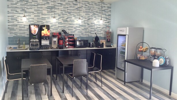 Cozinha moderna com painel de azulejos listrados, máquina de café, micro-ondas e geladeira. Há cadeiras pretas e uma mesa pequena.
