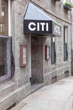 Um hotel chamado 'CITI HOTEL', com fachada de pedra, janelas grandes com grades de metal e marquise preta.