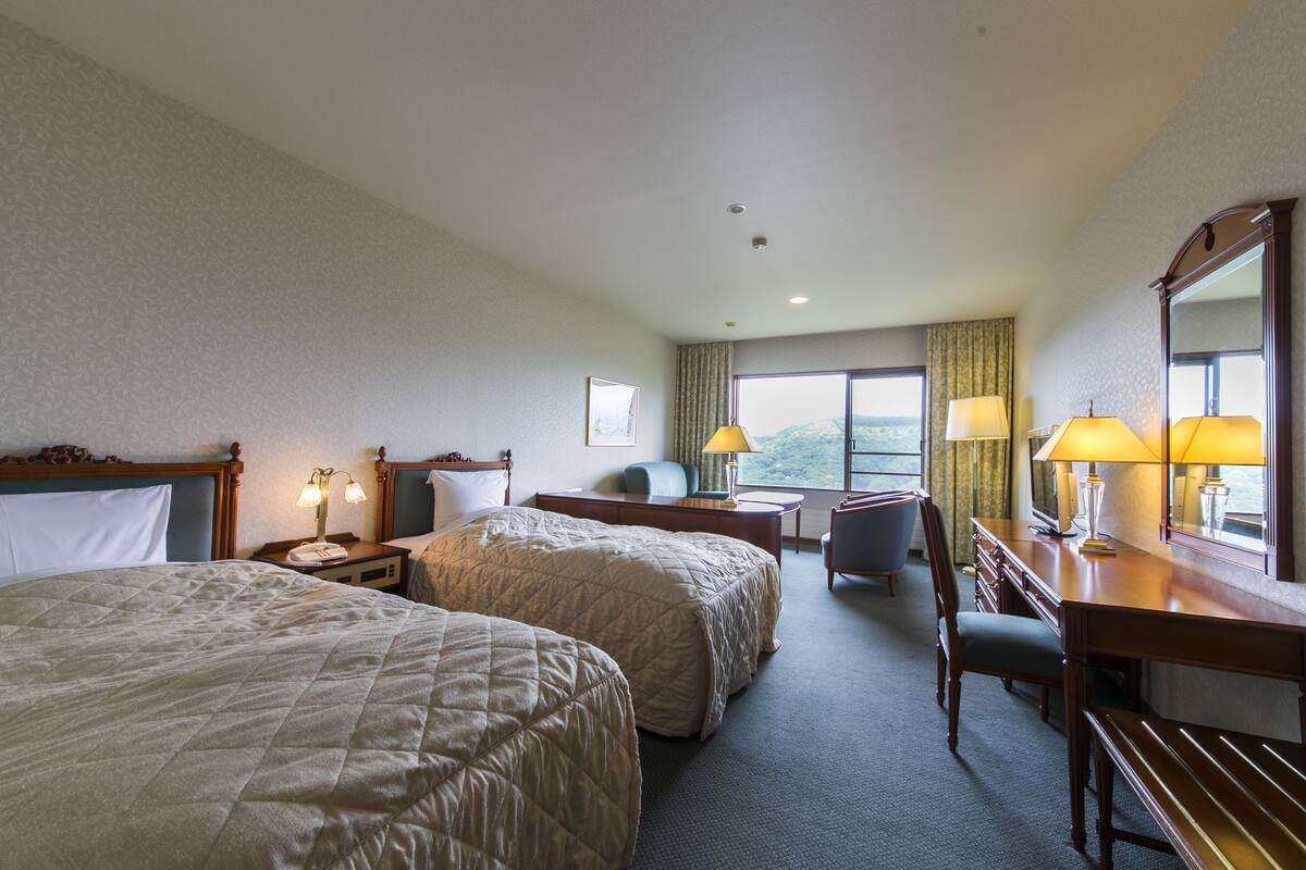 Quarto de hotel com duas camas, uma escrivaninha, uma cadeira, um abajur e uma janela grande.