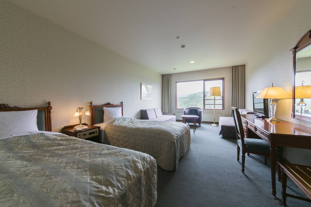 Quarto de hotel com duas camas, uma mesa, uma cadeira, uma televisão e uma janela com vista.